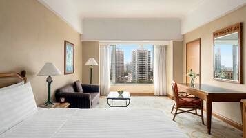 Quarto de hotel com uma cama grande, uma escrivaninha, uma cadeira, uma mesinha e vista para edifícios altos.