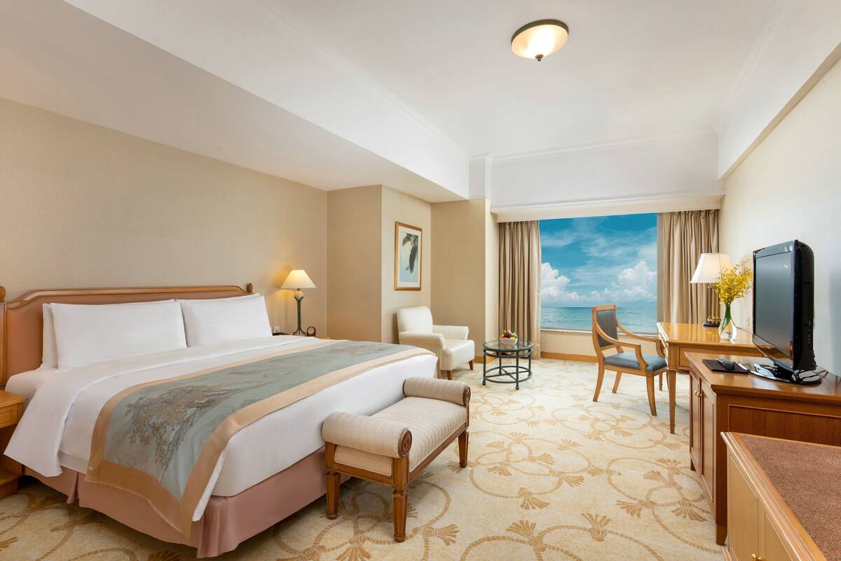 Quarto de hotel com uma cama grande, uma escrivaninha com computador, uma cadeira e vista para o oceano.