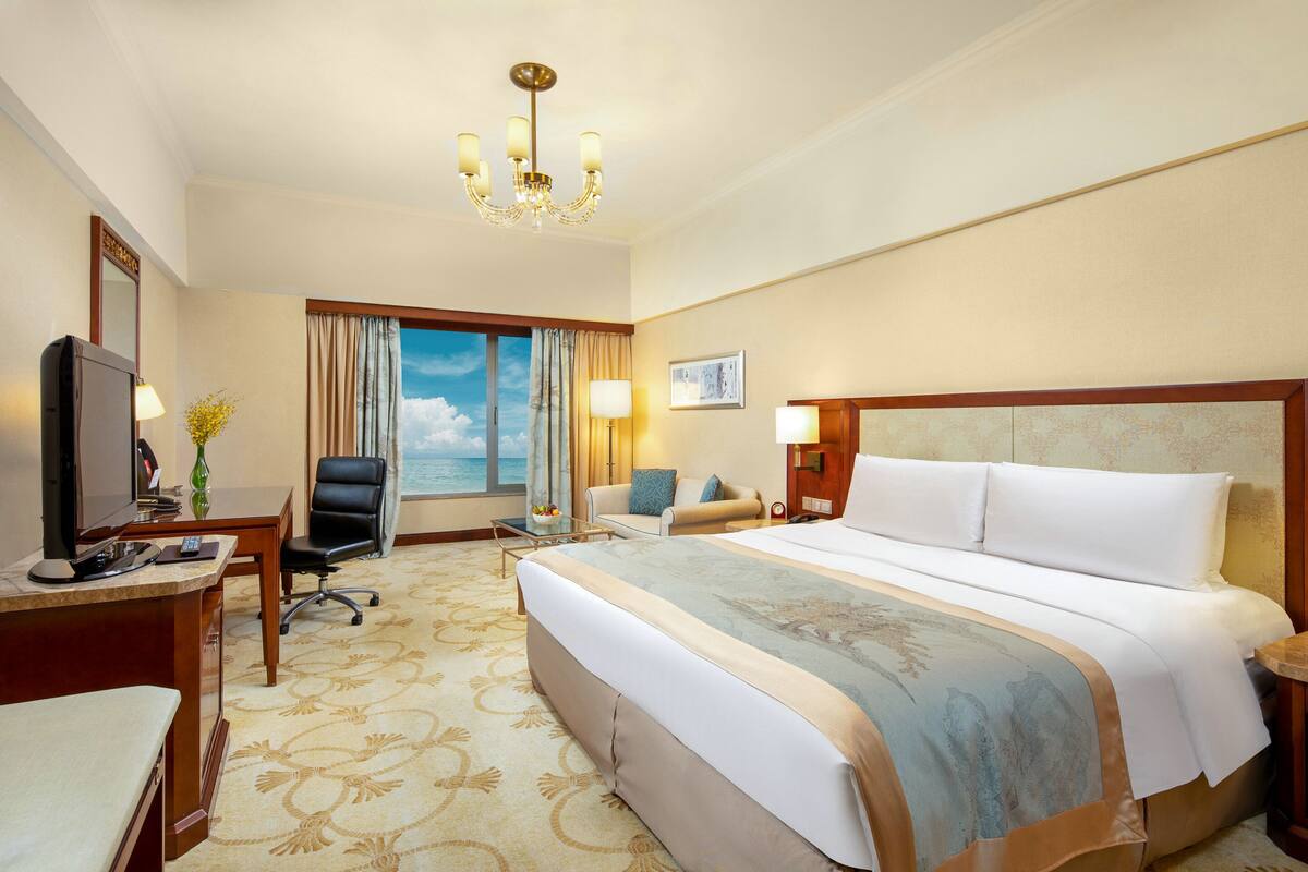 Quarto de hotel com uma cama grande, uma escrivaninha com computador, uma cadeira, uma televisão e uma janela com vista para o mar.