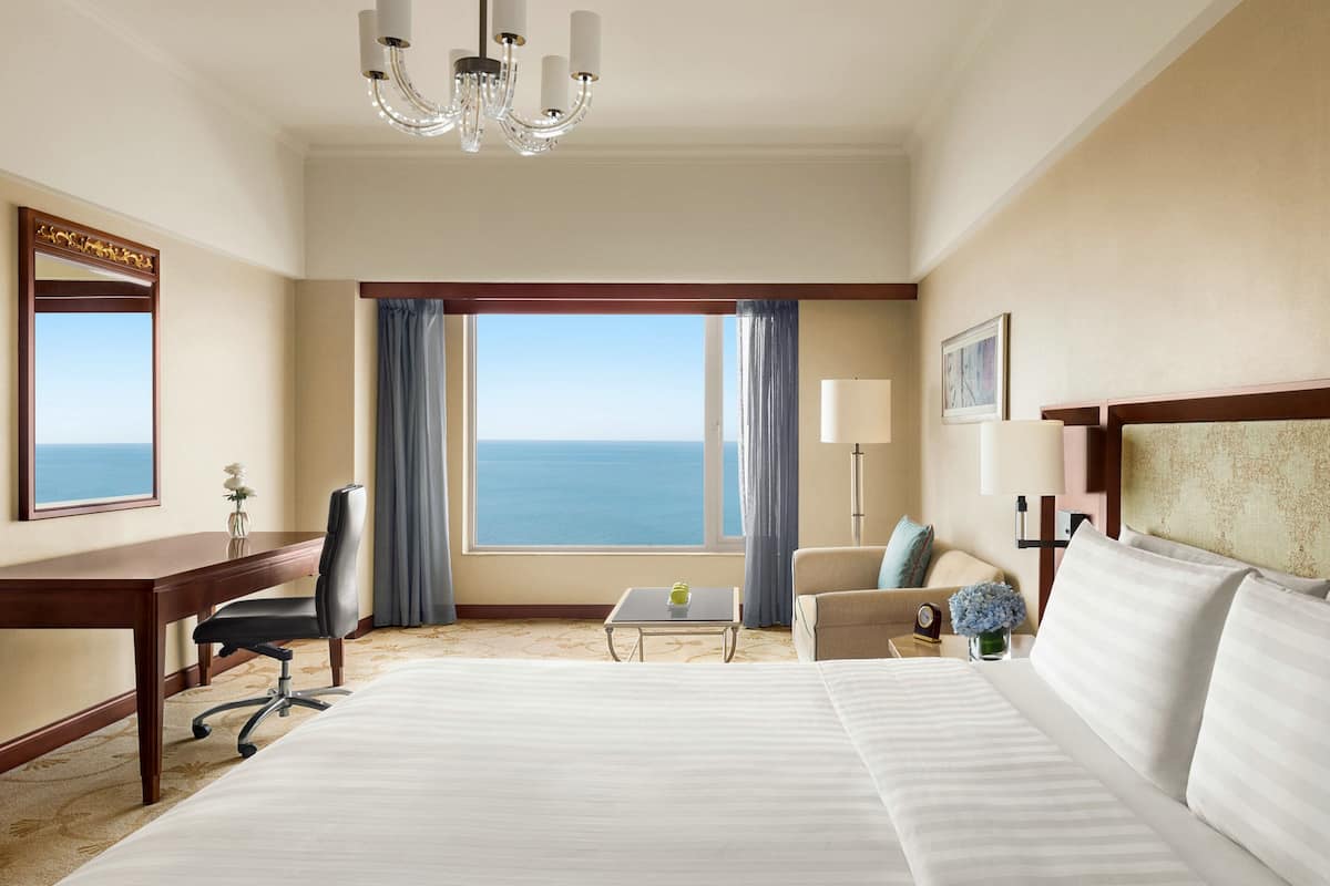 Quarto de hotel com uma cama grande, uma escrivaninha, uma cadeira, uma mesinha, uma luminária e vista para o oceano.