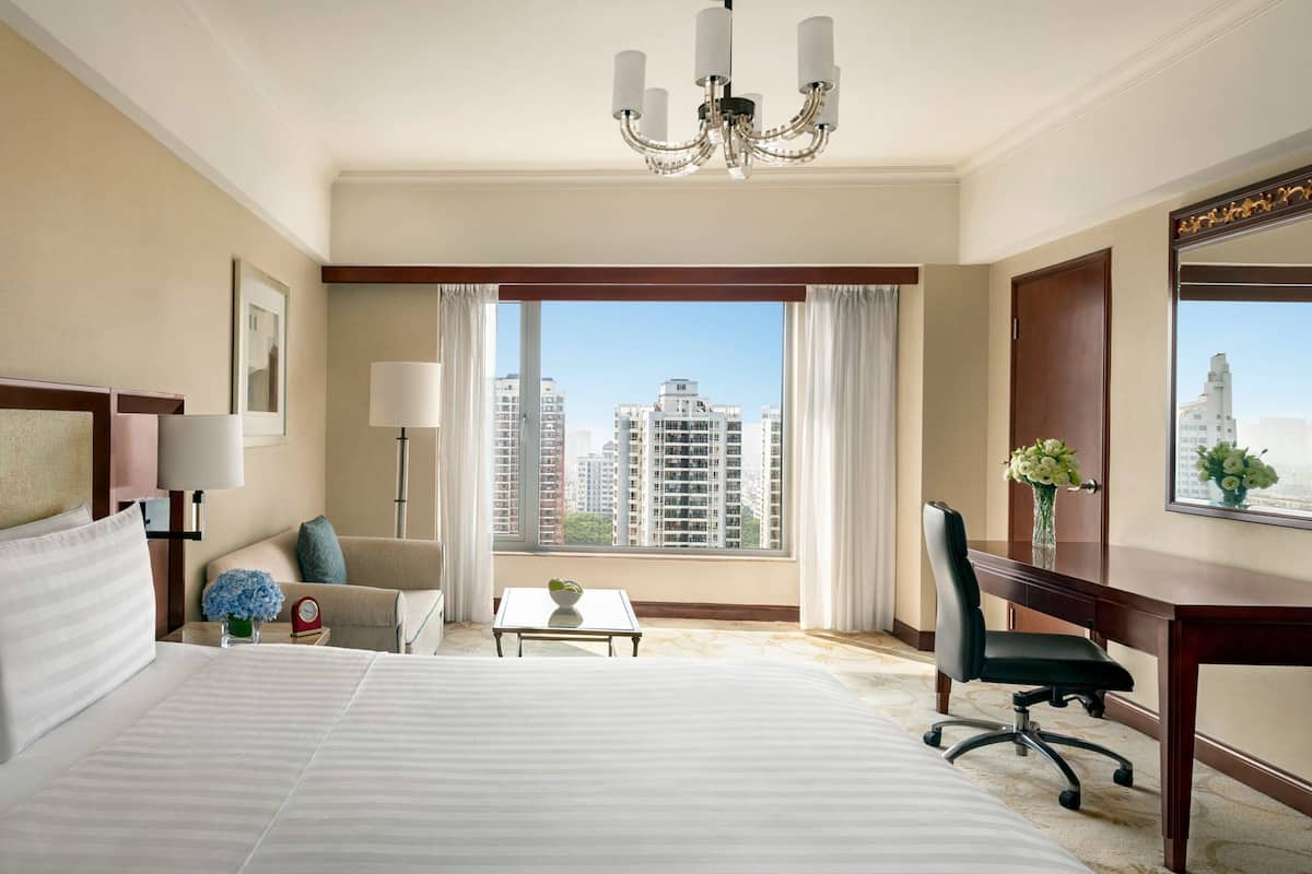 Quarto de hotel com uma cama grande, uma escrivaninha, uma cadeira, uma mesinha de centro com um vaso, e vista para edifícios altos pelas janelas amplas.
