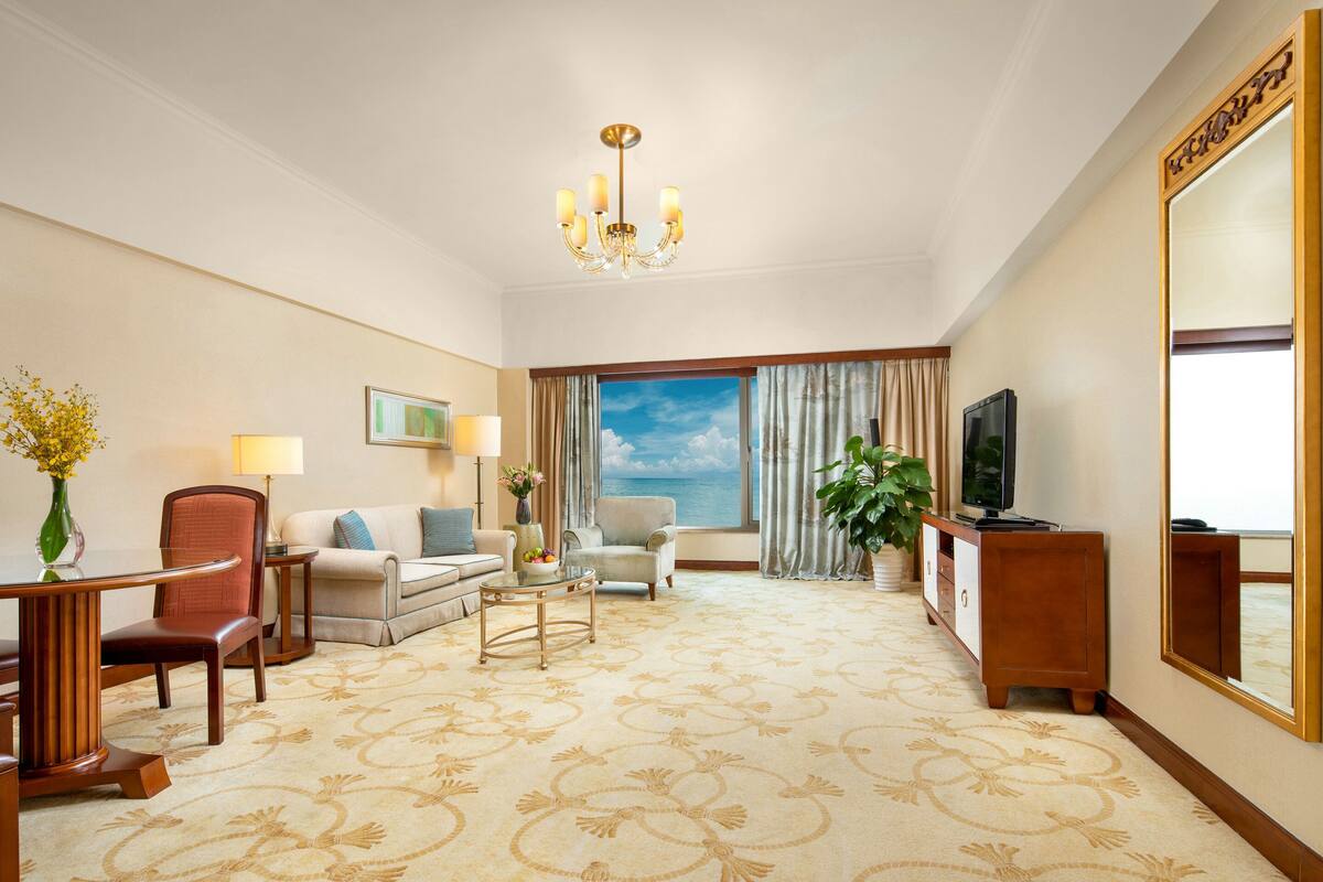 Quarto de hotel com uma grande janela, um lustre, uma televisão e um espaço de jantar com mesa e cadeiras.