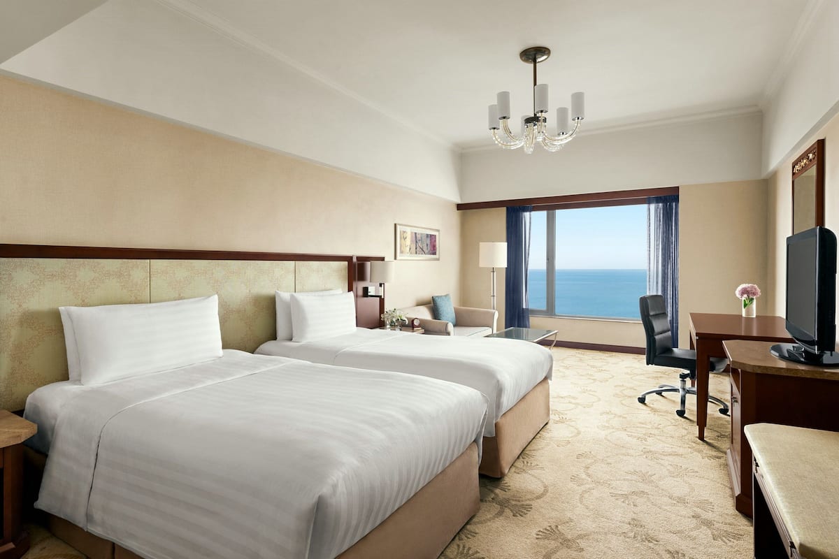 Quarto de hotel com duas camas, uma escrivaninha, uma cadeira, televisão e uma janela grande com vista para o mar.