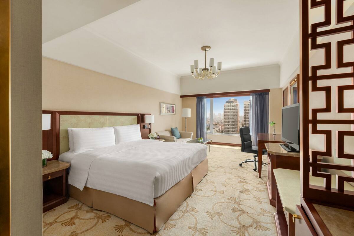 Quarto de hotel com uma cama grande, uma escrivaninha com computador, uma cadeira, uma televisão e vista para edifícios altos.