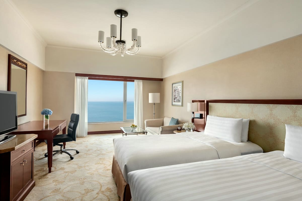 Quarto de hotel com duas camas, uma escrivaninha com cadeira, televisão e vista para o oceano.
