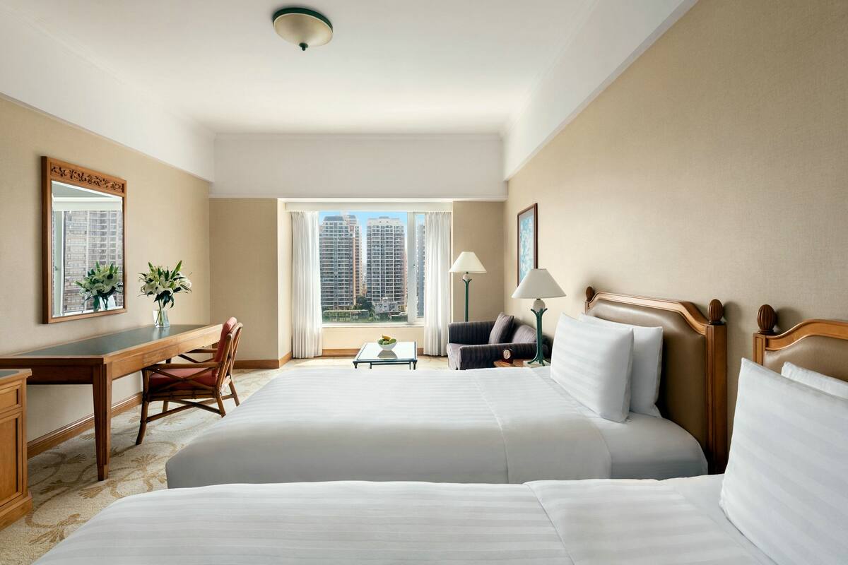 Quarto de hotel com duas camas, uma escrivaninha, uma cadeira, um abajur e vista para edifícios altos.