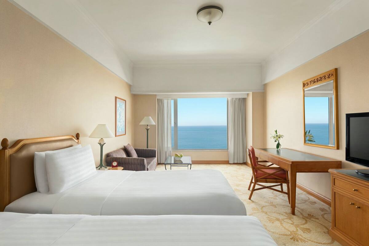 Quarto de hotel com duas camas, uma escrivaninha, uma cadeira, uma televisão e uma janela grande com vista para o mar.