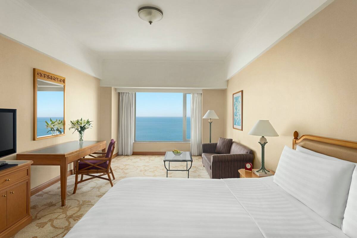 Quarto de hotel com uma cama grande, uma escrivaninha, uma cadeira, uma televisão, um abajur e vista para o mar.
