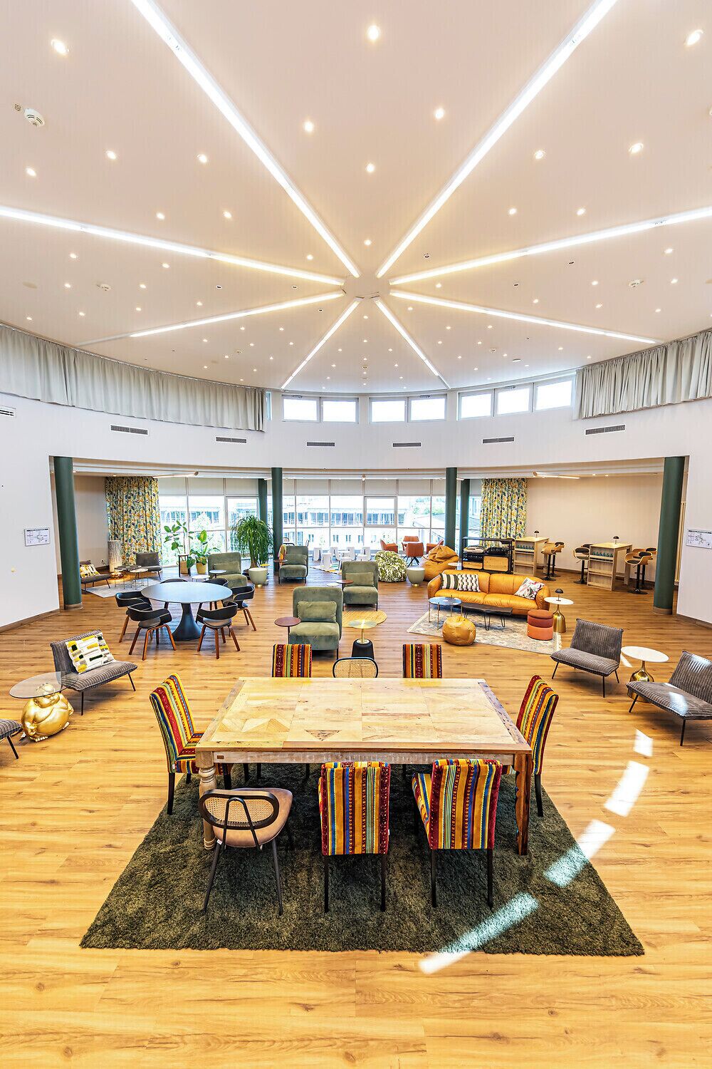 Amplo espaço interno com mesa redonda central rodeada por cadeiras coloridas, teto alto com iluminação em formato de estrela e vista para o exterior por grandes janelas.