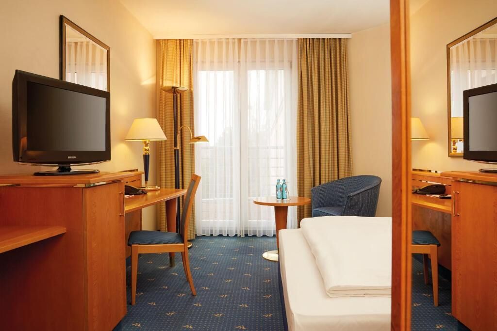 Quarto de hotel com cama, escrivaninha com televisão, cadeira, mesinha com abajur e janela com cortinas.