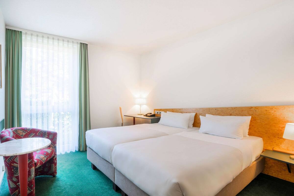 Quarto de hotel com duas camas, uma mesinha, uma cadeira e um abajur.