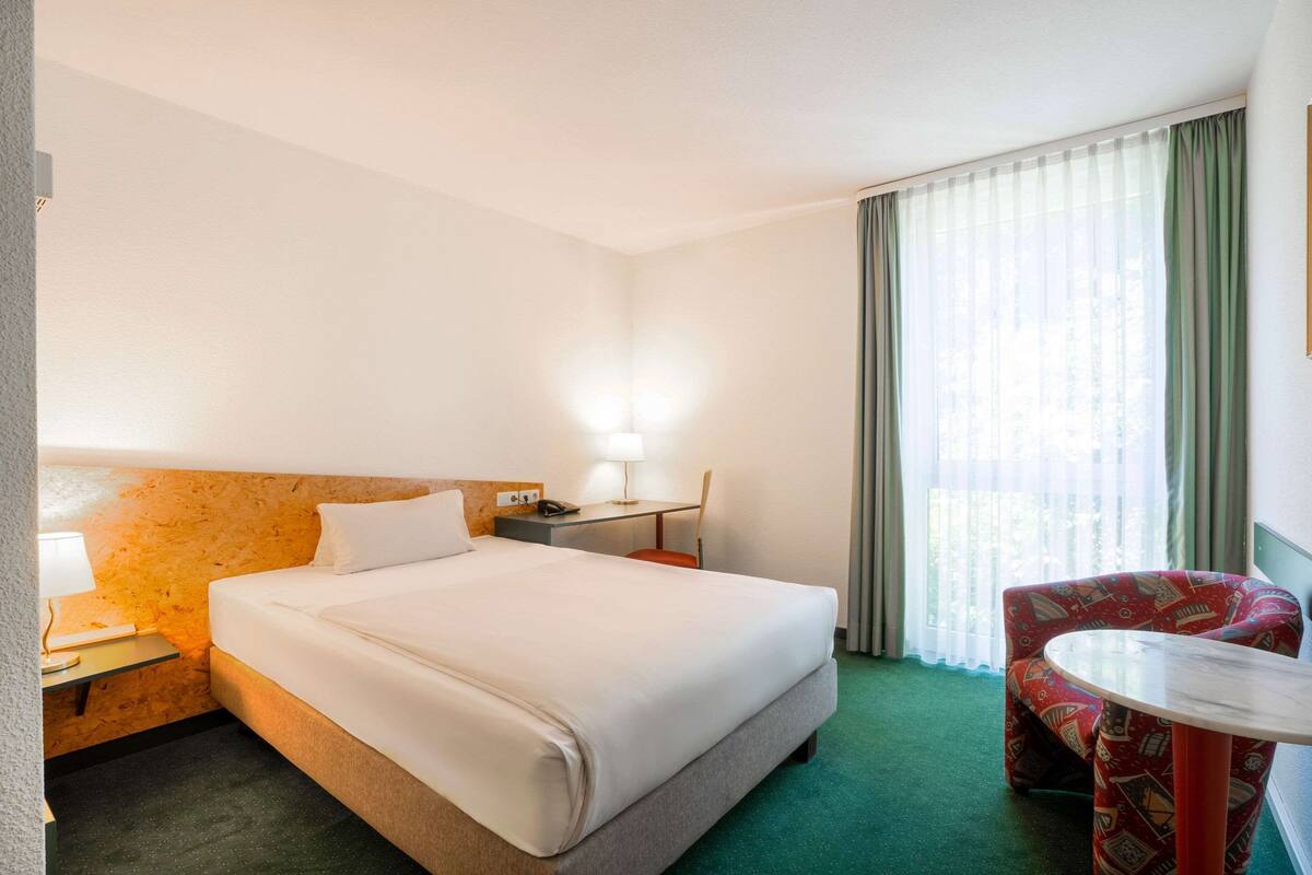 Quarto de hotel com uma cama grande, uma escrivaninha com abajur, uma cadeira e uma janela com cortinas.