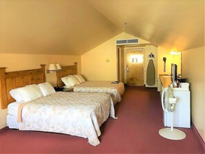 Quarto de hotel com duas camas, uma escrivaninha, uma cadeira, um espelho, um ventilador e uma porta para o banheiro.