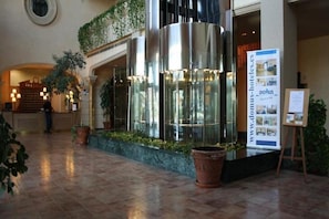 Um saguão de hotel com um elevador central, um balcão de recepção e uma escadaria.
