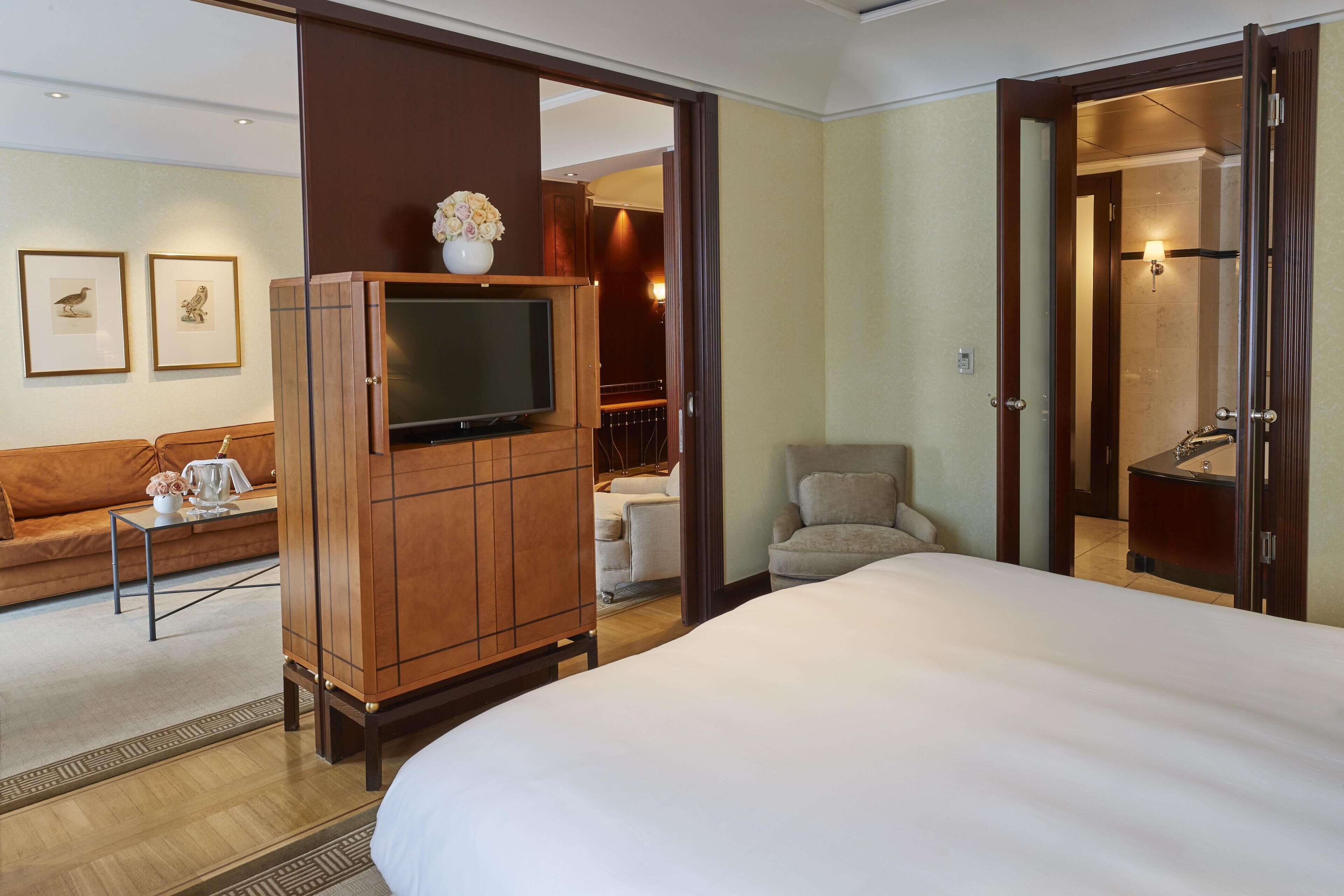 Quarto de hotel com cama, televisão em um móvel de madeira, sofá, poltrona e banheiro visível por uma porta aberta.