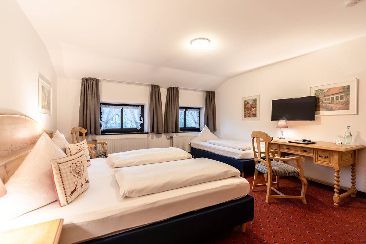 Quarto de hotel com cama, escrivaninha, cadeira, televisão e janelas com cortinas.
