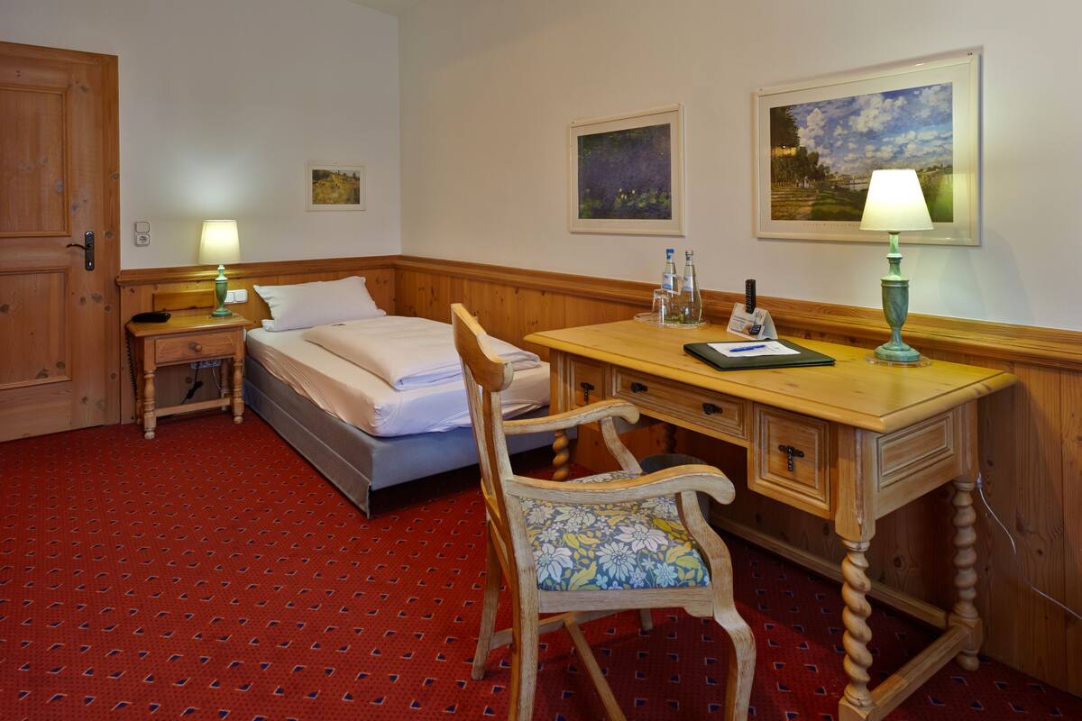 Quarto de hotel com cama, escrivaninha, cadeira e duas pinturas na parede.