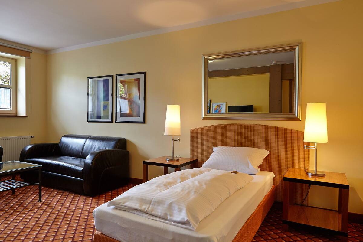 Quarto de hotel com cama, poltrona de couro, mesinha, duas luminárias de cabeceira e duas molduras com quadros na parede.