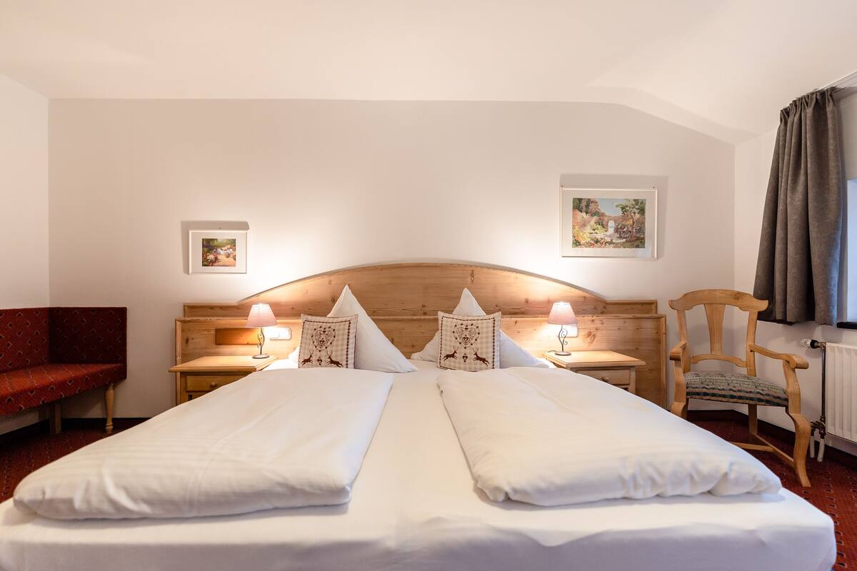 Uma cama de casal com duas almofadas, cabeceira de madeira, mesas de cabeceira com luminárias, uma cadeira e dois quadros na parede.