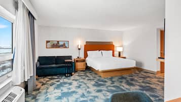 Quarto de hotel com cama, escrivaninha, poltrona, sofá, mesa de cabeceira e janela com cortinas.