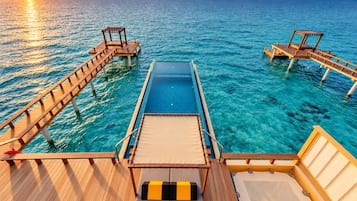 Um deck de madeira com piscina, cadeiras de descanso e vista para o oceano ao pôr do sol.