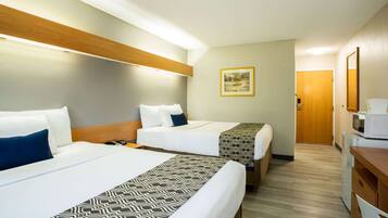 Quarto de hotel com duas camas, uma cabeceira de madeira, um quadro na parede e um micro-ondas sobre uma bancada de madeira.