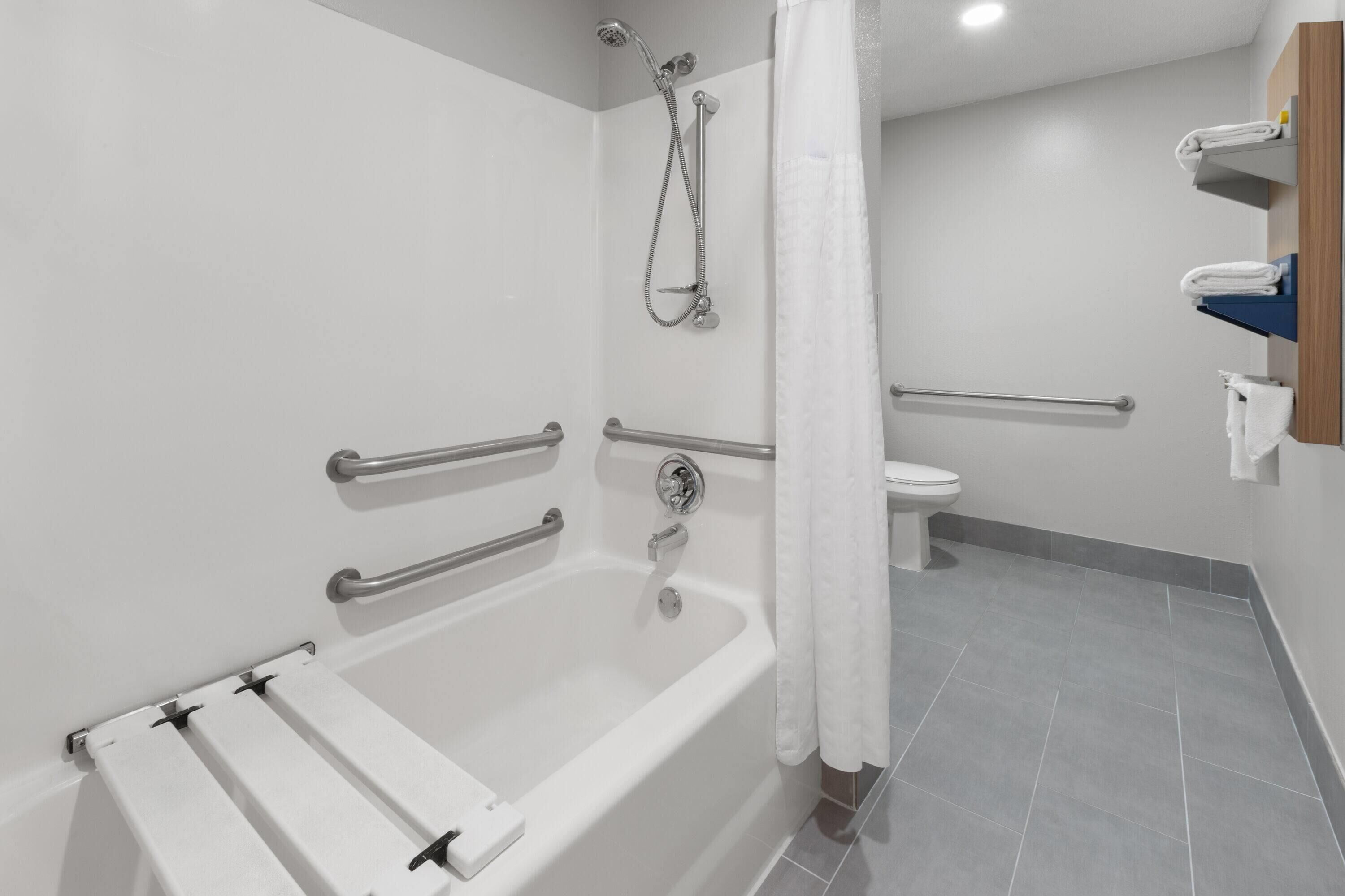 Casa de banho moderna com banheira, chuveiro e sanita. Inclui barras de apoio e porta-toalhas.