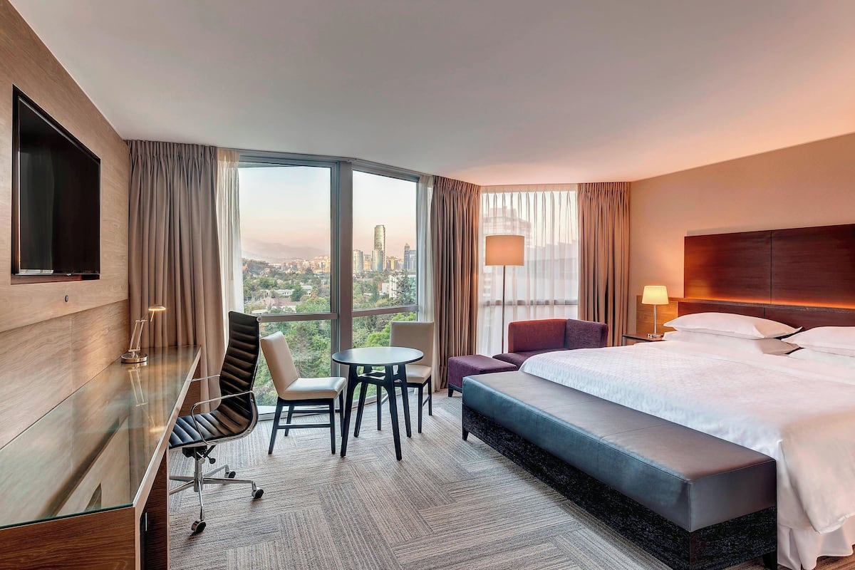 Quarto de hotel com uma cama grande, uma escrivaninha com televisão, uma cadeira, uma mesinha e vista para a cidade através de amplas janelas.