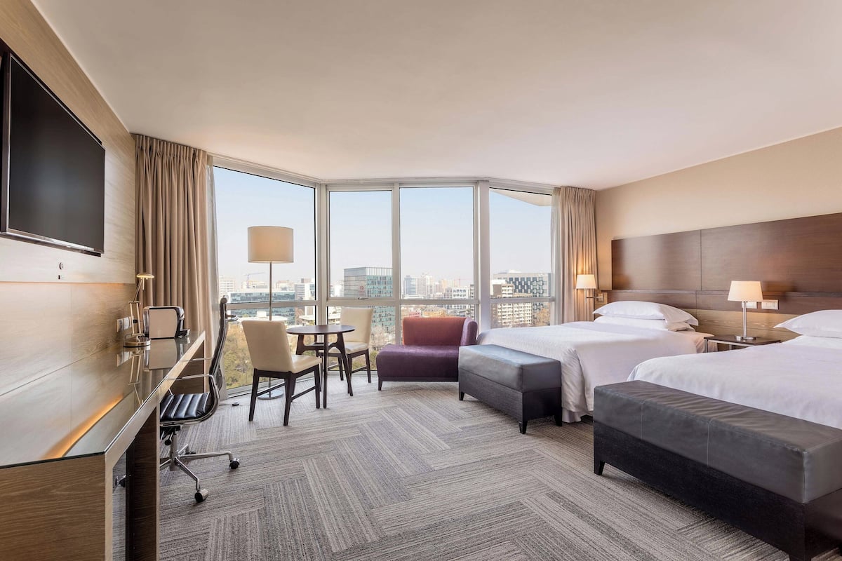 Quarto de hotel com uma janela grande, uma escrivaninha, duas camas e uma área de estar.