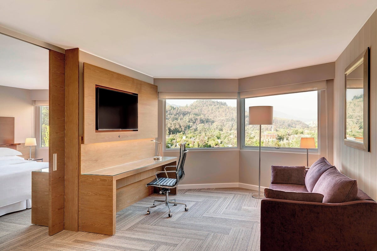 Quarto de hotel com cama, escrivaninha com televisão, sofá e uma grande janela com vista para as montanhas.