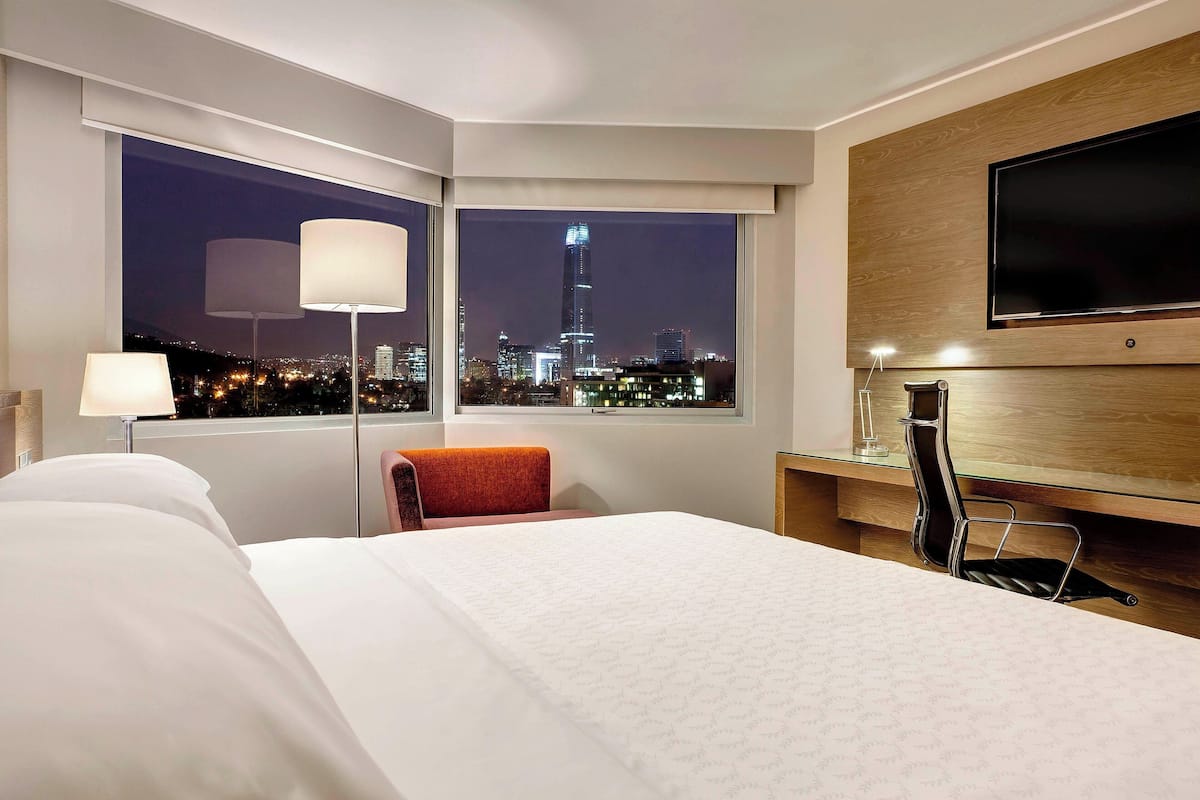 Quarto de hotel com cama, uma cadeira, uma escrivaninha, televisão e uma janela grande com vista para a cidade à noite.