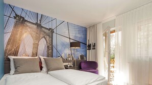 Quarto de hotel com uma cama grande, uma cadeira roxa, um mural com paisagem urbana e vista para o exterior.