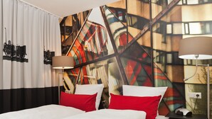 Quarto de hotel com cama, duas almofadas vermelhas, abajur e parede com pintura abstrata colorida.