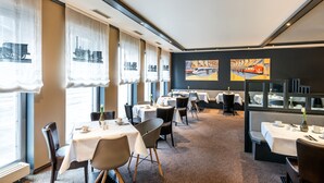 Uma área de jantar de hotel com mesas preparadas para o serviço, grandes janelas com cortinas e obras de arte nas paredes.