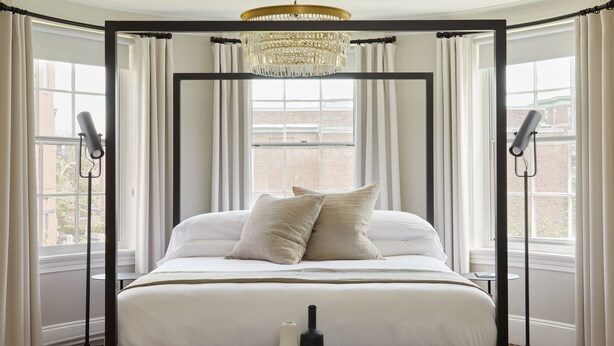 Uma cama com dossel e lustre, uma mesinha de cabeceira com livros, e vista para um prédio de tijolos pelas janelas.
