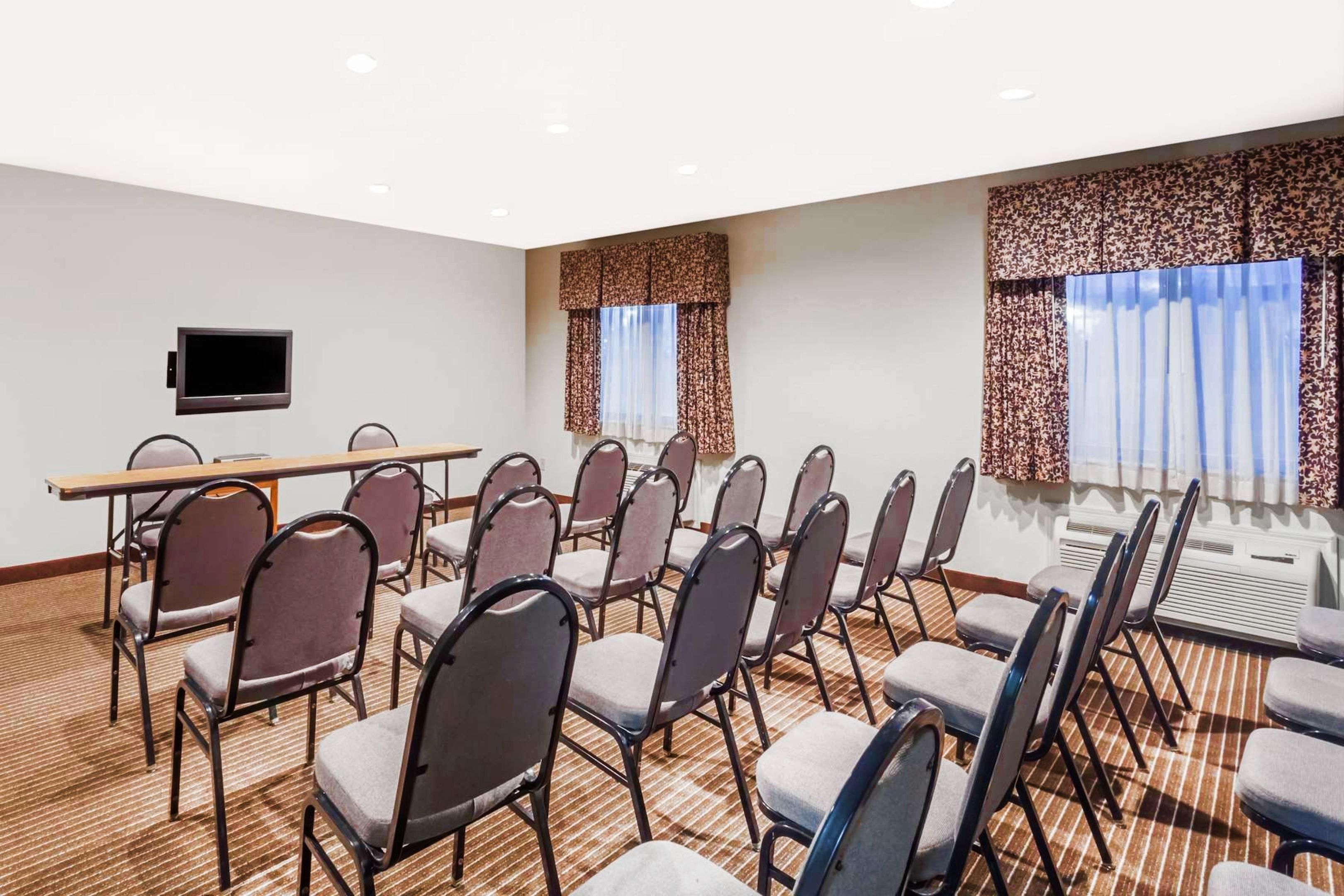 Uma sala de conferências com fileiras de cadeiras, uma televisão e janelas com cortinas.