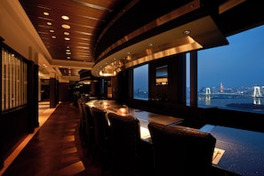 Um restaurante com vista para o horizonte urbano, apresentando um balcão comprido com cadeiras e um teto alto com iluminação embutida.