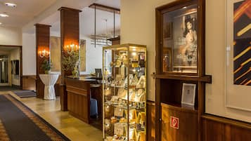 Um saguão de hotel com balcão de recepção, uma vitrine de vidro com joias e obras de arte emolduradas nas paredes.