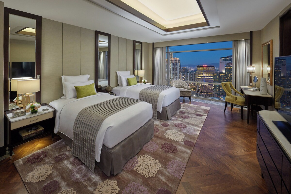 Quarto de hotel com duas camas, uma mesa, uma cadeira e uma janela grande com vista para a cidade.