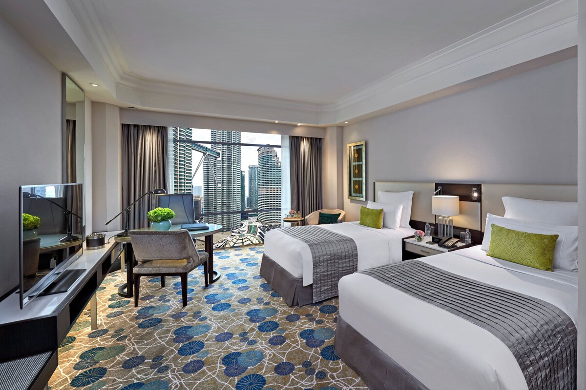 Quarto de hotel com uma cama grande, uma escrivaninha com computador, uma cadeira, uma mesinha de cabeceira com abajur e vista para prédios altos.
