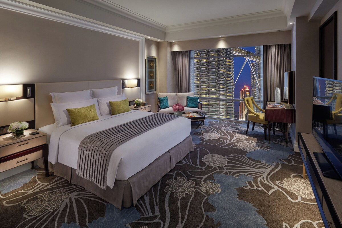 Quarto de hotel com uma cama grande, uma mesa, uma cadeira e uma televisão. Há vista para a cidade pela janela.
