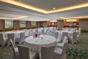 Um amplo salão de banquetes com várias mesas redondas, cada uma com toalhas brancas e cadeiras, e um grande candelabro ao centro.