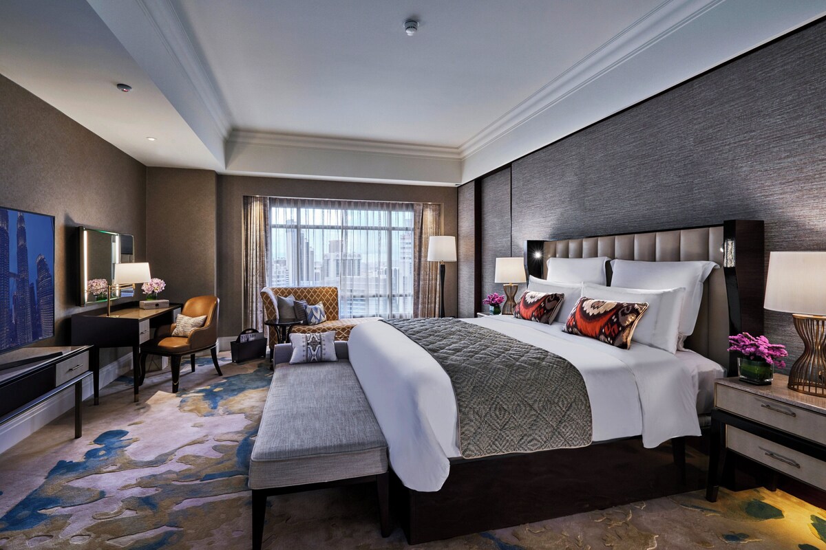 Um quarto de hotel espaçoso com uma cama grande, uma escrivaninha, uma cadeira, uma televisão e uma janela ampla com cortinas.