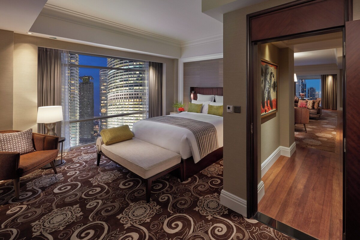 Quarto de hotel com uma cama grande, área de estar com uma poltrona e uma mesa, vista para a cidade através de grandes janelas e um tapete estampado.