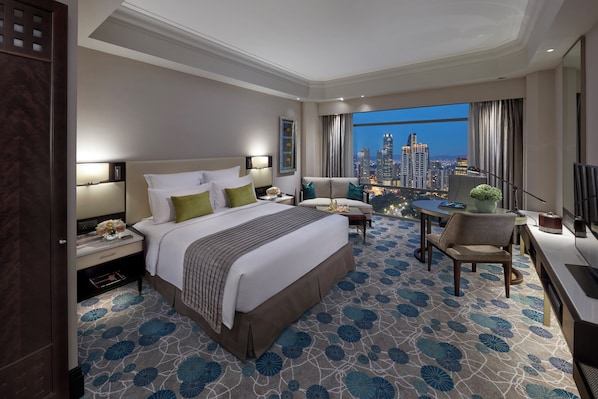 Quarto de hotel com uma cama grande, uma escrivaninha, uma cadeira, uma mesinha e vista para a paisagem urbana.