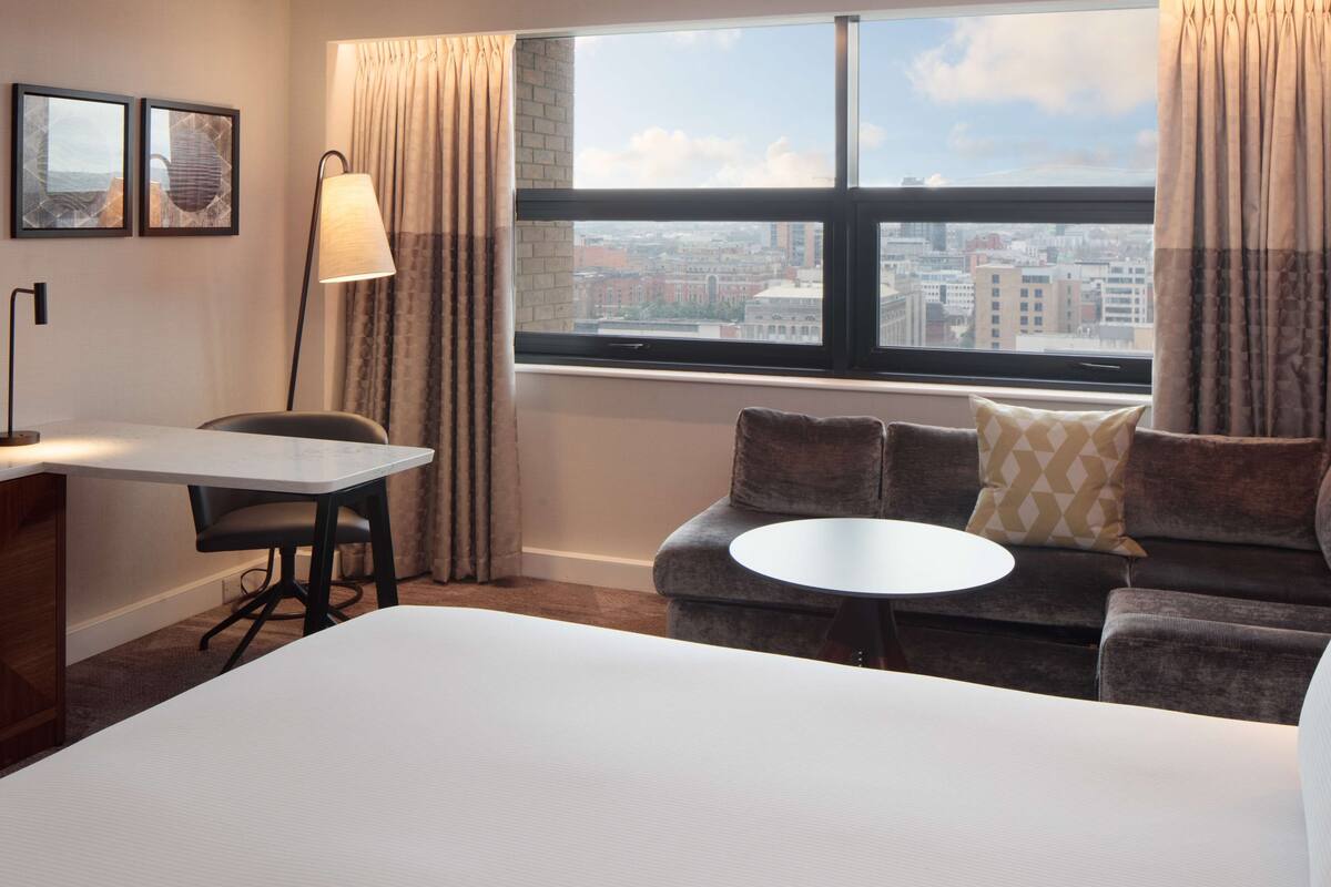 Quarto de hotel com cama, escrivaninha, cadeira, sofá e vista da cidade através de amplas janelas.