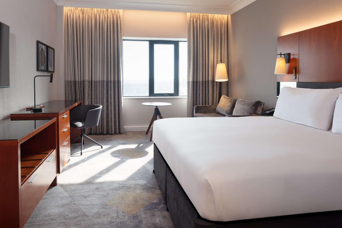 Quarto de hotel com uma cama grande, uma escrivaninha com cadeira, uma mesa redonda pequena e uma luminária.