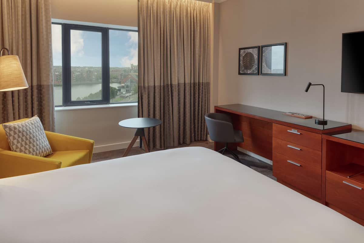 Quarto de hotel com uma cama grande, uma escrivaninha, uma cadeira, uma mesa pequena e vista para a paisagem urbana através de uma janela.