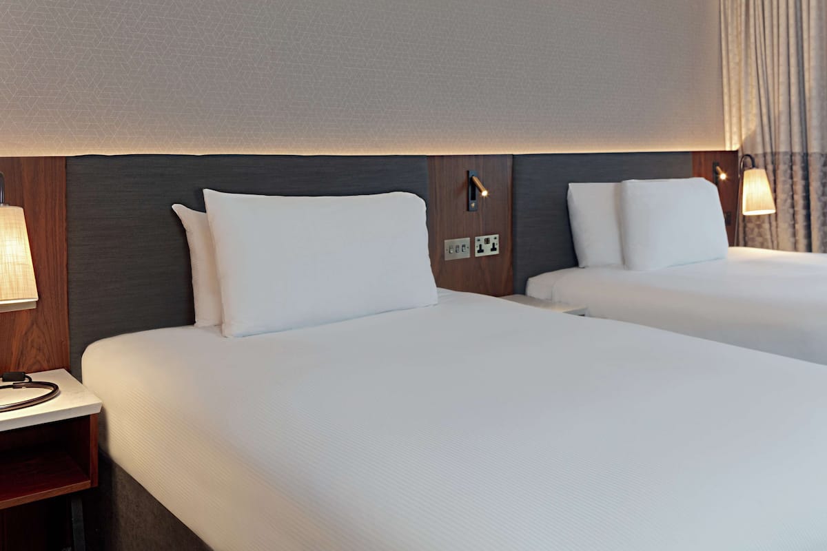 Quarto de hotel com duas camas, uma mesa de cabeceira e uma luminária.