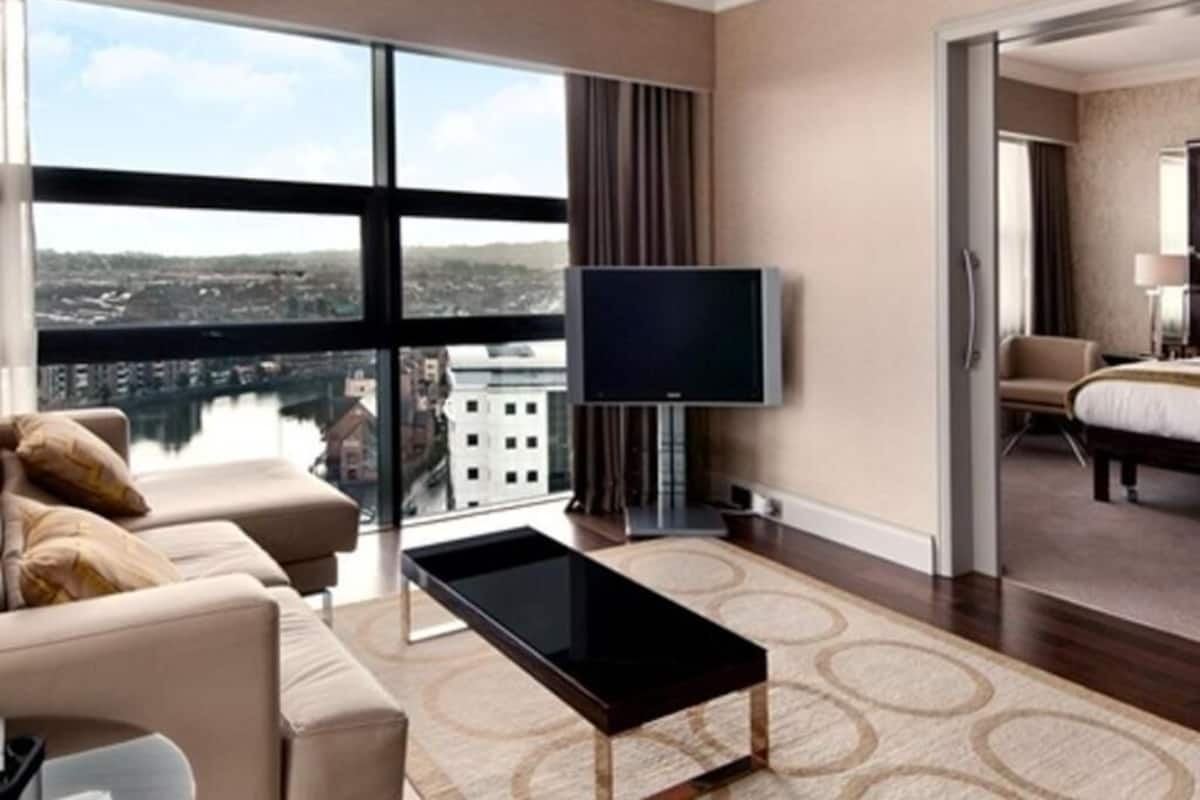 Quarto de hotel moderno com uma janela grande, televisão de tela plana, sofá, mesa de centro e uma cama.